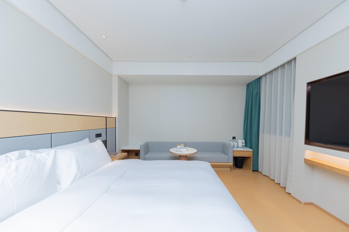 Quarto de hotel com uma cama grande, área de estar com uma mesa e uma televisão na parede.
