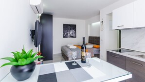 Quarto de hotel moderno com cama, área de refeições com mesa e cadeiras, cozinha e planta em vaso.