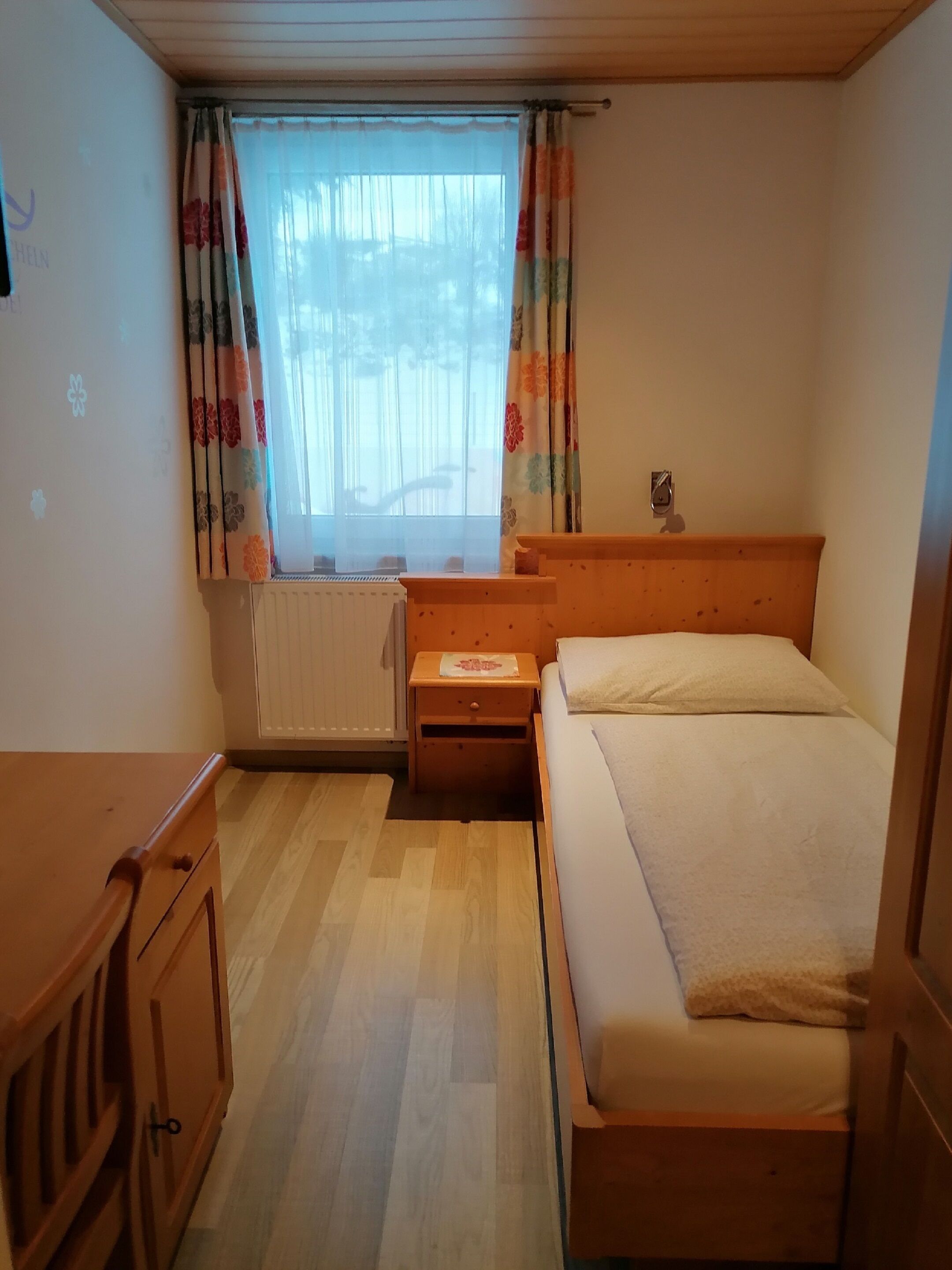 Quarto compacto com uma cama de solteiro, mesa de cabeceira, cômoda de madeira e janela com cortinas florais.