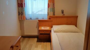 Quarto compacto com uma cama de solteiro, mesa de cabeceira, cômoda de madeira e janela com cortinas florais.