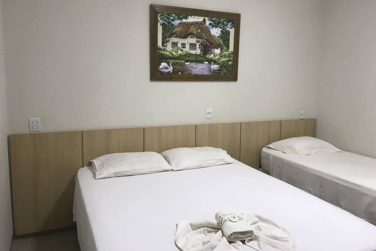 Quarto de hotel com duas camas de solteiro, cabeceira de madeira e um quadro de uma casa de campo na parede.