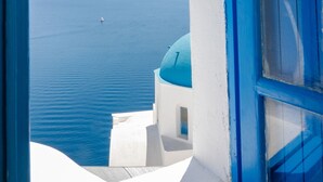 Vista através de uma janela emoldurada por azul de um prédio branco com cúpula azul, sobre um mar calmo.