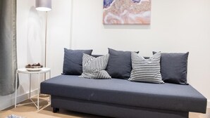Uma sala de estar moderna com um sofá cinza escuro, duas almofadas estampadas em preto e branco, uma mesinha lateral branca com uma tigela de pinhas, e um abajur.