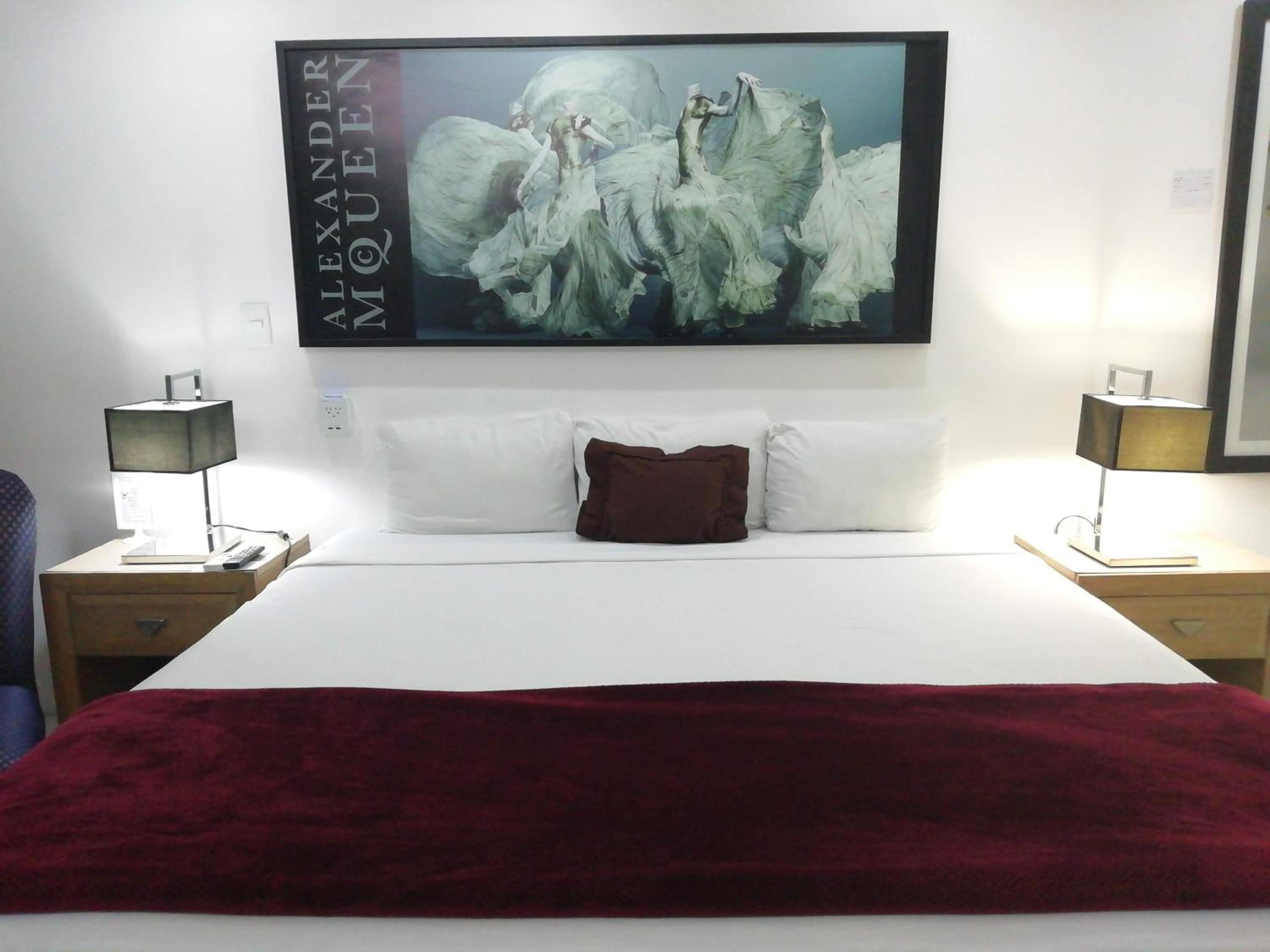 Quarto de hotel com uma grande pintura de uma escultura, uma cama branca com manta vermelha, duas luminárias de cabeceira e uma mesinha de noite com um livro e controle remoto.