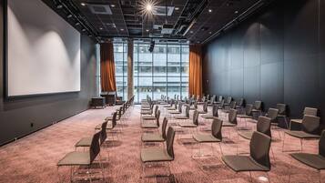 Uma sala de conferências com fileiras de cadeiras, uma tela grande e vista para um edifício através de amplas janelas.