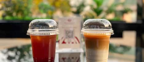 Duas bebidas geladas em copos de plástico transparente sobre uma mesa de vidro, com uma vista desfocada do exterior ao fundo.