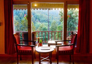 Quarto com duas cadeiras e uma mesa, janela com vista para uma floresta e porta com vista para uma varanda.