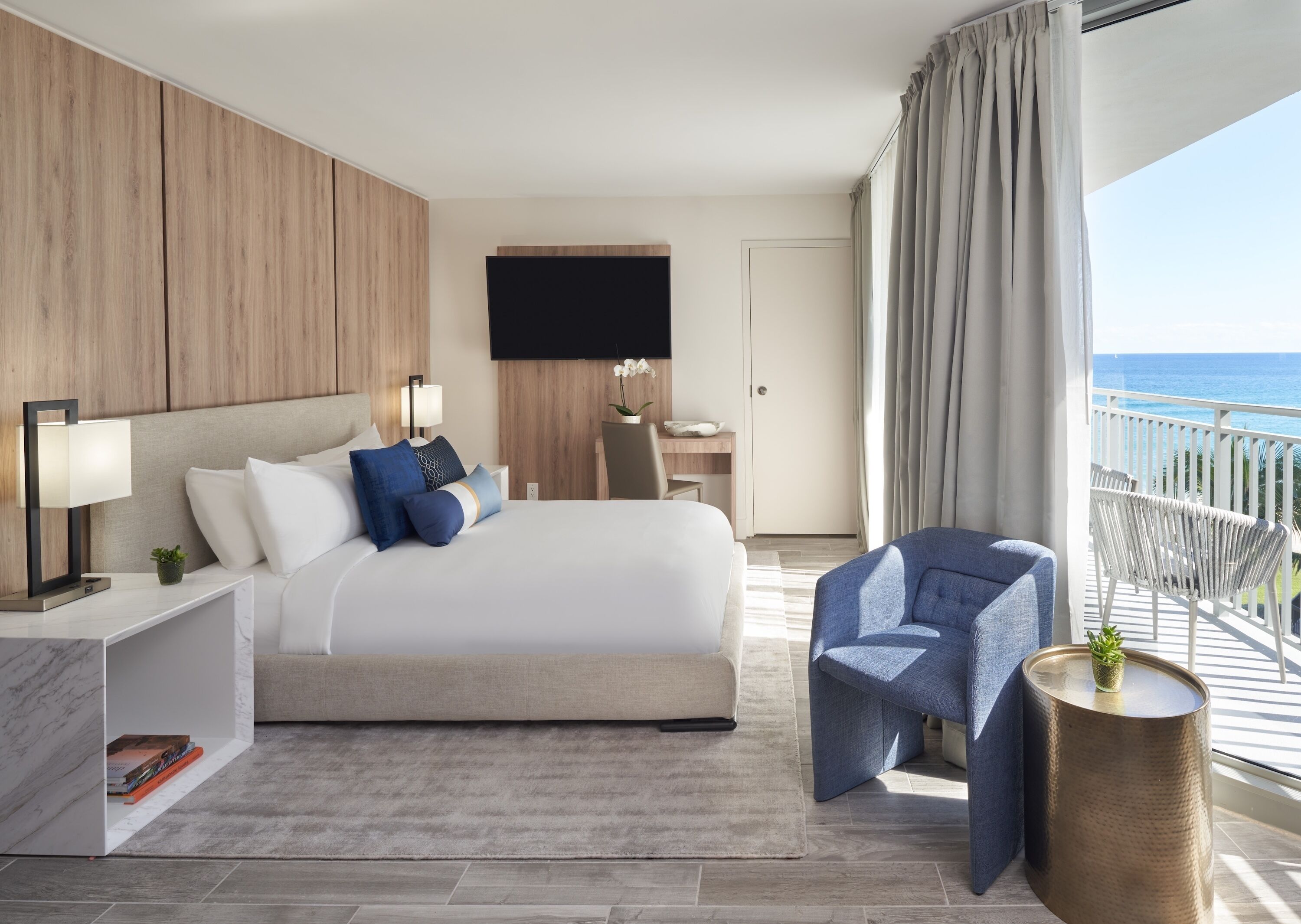 Quarto de hotel com cama, mesa de cabeceira, abajur, cadeira, escrivaninha, televisão e varanda com vista para o oceano.