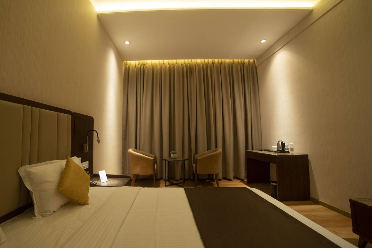Quarto de hotel com cama, duas cadeiras, uma mesinha, escrivaninha e cortina.