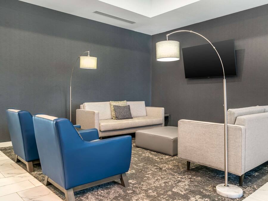 Uma sala de estar moderna com um sofá cinza, poltronas azuis e um banquinho cinza. Uma luminária de chão e uma TV de tela plana estão fixadas na parede.