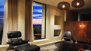Uma sala de estar moderna com um sofá, uma mesa de jantar e duas cadeiras. Há uma janela grande com cortinas, vista do céu e uma parede de madeira com luminária.