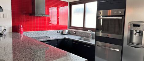 Cozinha moderna com bancadas de granito, eletrodomésticos de aço inoxidável e painel de azulejos vermelhos.