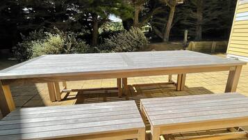 Mesa de madeira com bancos em um pátio, cercado por um jardim com árvores e arbustos.