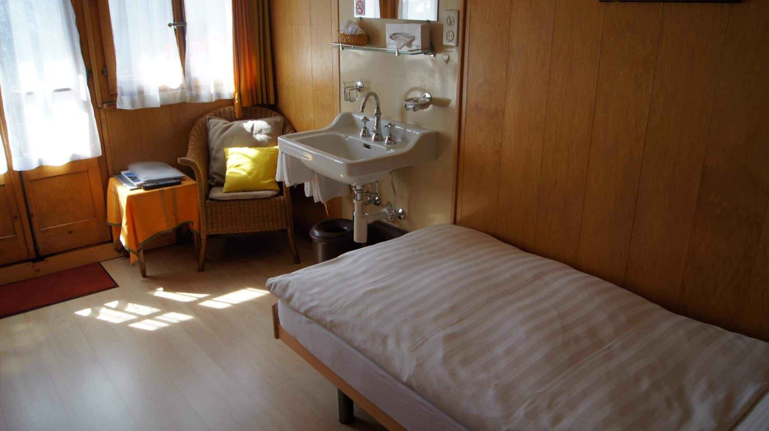 Quarto pequeno e acolhedor com uma cama de solteiro, guarda-roupa de madeira, cadeira de vime com almofada amarela, uma mesa pequena com um controle remoto e uma pia branca com espelho acima.