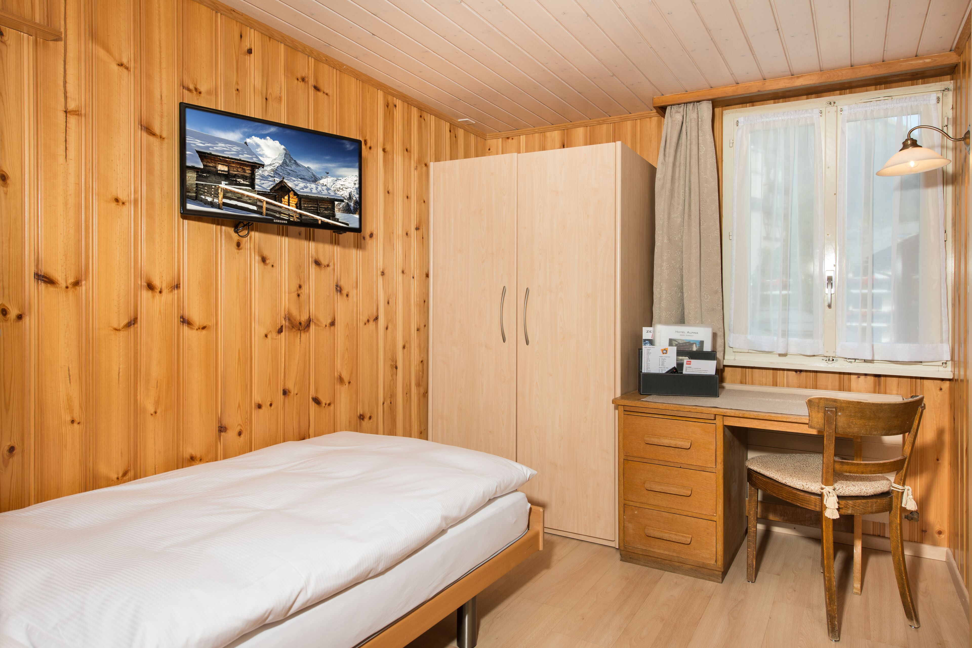 Quarto de madeira com cama, escrivaninha, cadeira, guarda-roupa e janela com cortinas.