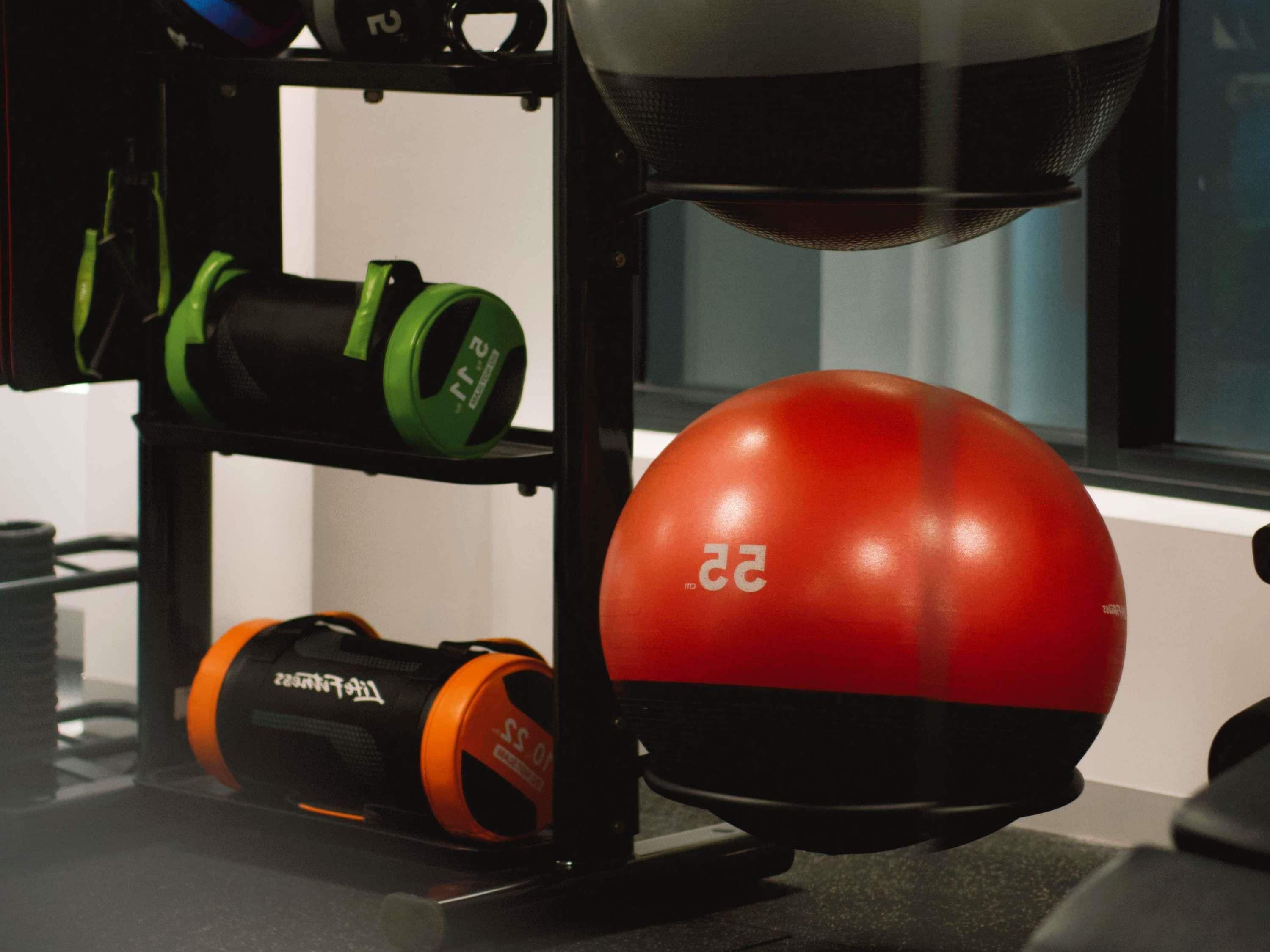 Uma academia com uma bola de exercício vermelha, equipamentos de musculação verde e laranja, e uma janela ao fundo.