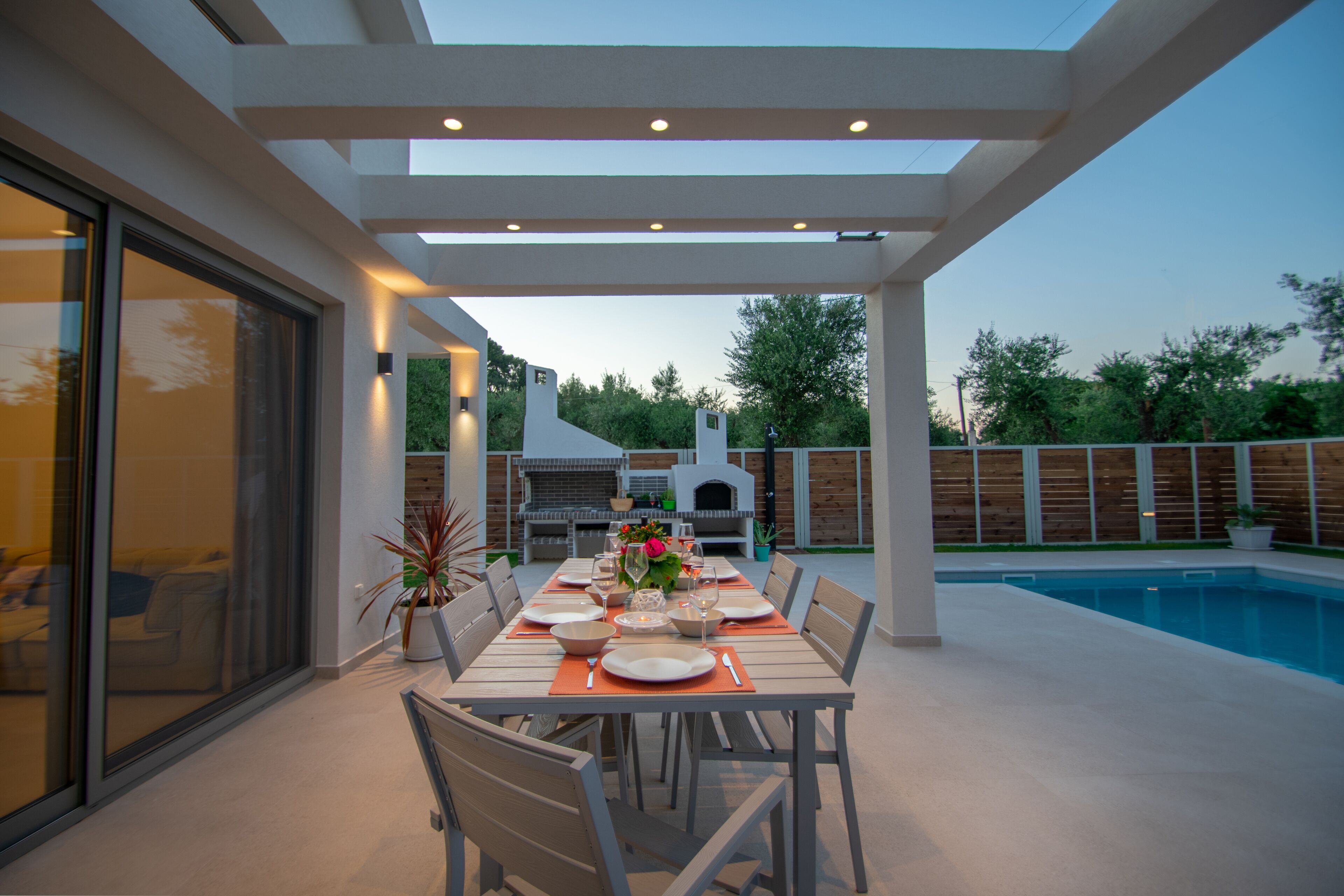 Um pátio externo com uma mesa de jantar para seis pessoas, rodeada por cadeiras. Há uma piscina ao fundo e um forno de tijolos à esquerda.