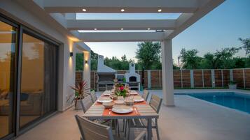 Um pátio externo com uma mesa de jantar para seis pessoas, rodeada por cadeiras. Há uma piscina ao fundo e um forno de tijolos à esquerda.