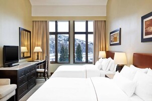 Quarto de hotel com duas camas, uma escrivaninha, televisão e amplas janelas com vista para uma montanha coberta de neve.