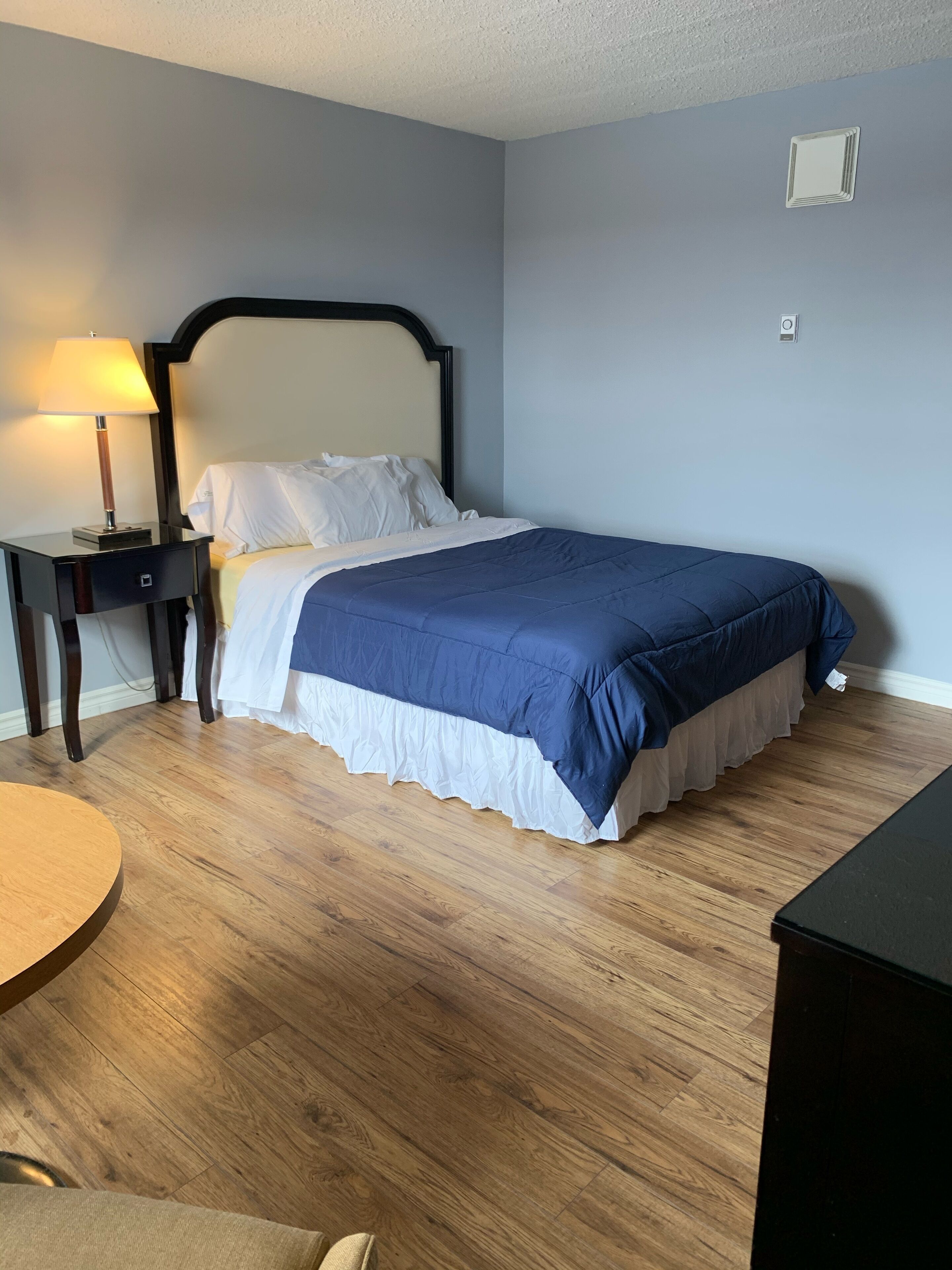 Cama bem arrumada com edredom azul e branco, luminária de cabeceira e piso de madeira.