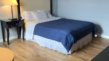 Cama bem arrumada com edredom azul e branco, luminária de cabeceira e piso de madeira.