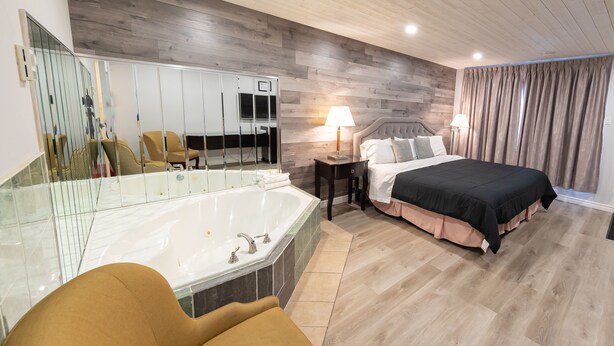 Quarto de hotel moderno com uma banheira grande, uma cama com roupa de cama preta, uma mesa de cabeceira com abajur e um espelho grande.