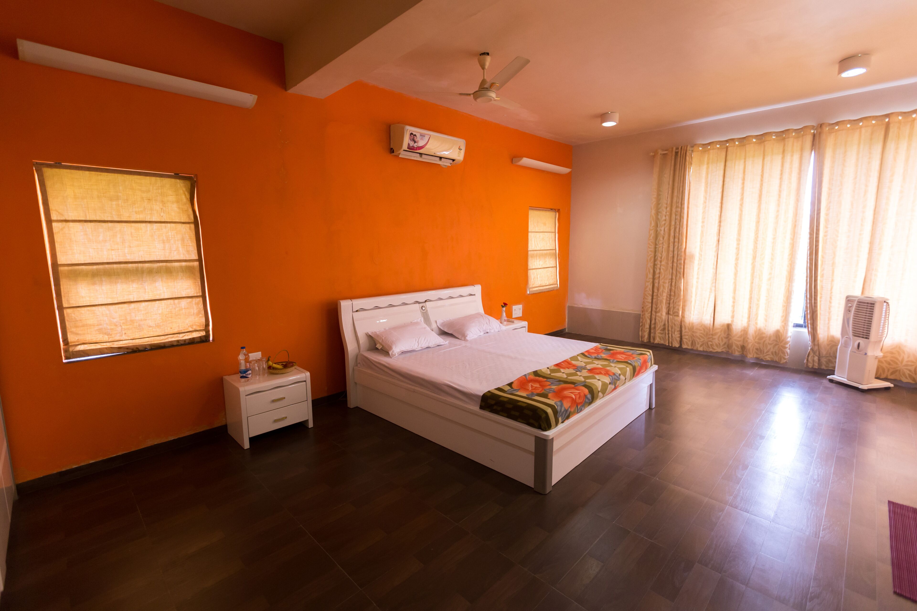 Quarto com parede laranja, cama com roupa de cama branca, mesa de cabeceira, janela com persianas e ar condicionado.