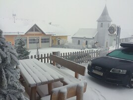 Uma área externa coberta de neve com um banco e uma mesa, um carro estacionado por perto, e um edifício com uma placa indicando "FEUERWEHR BRAUNEGG" ao fundo.