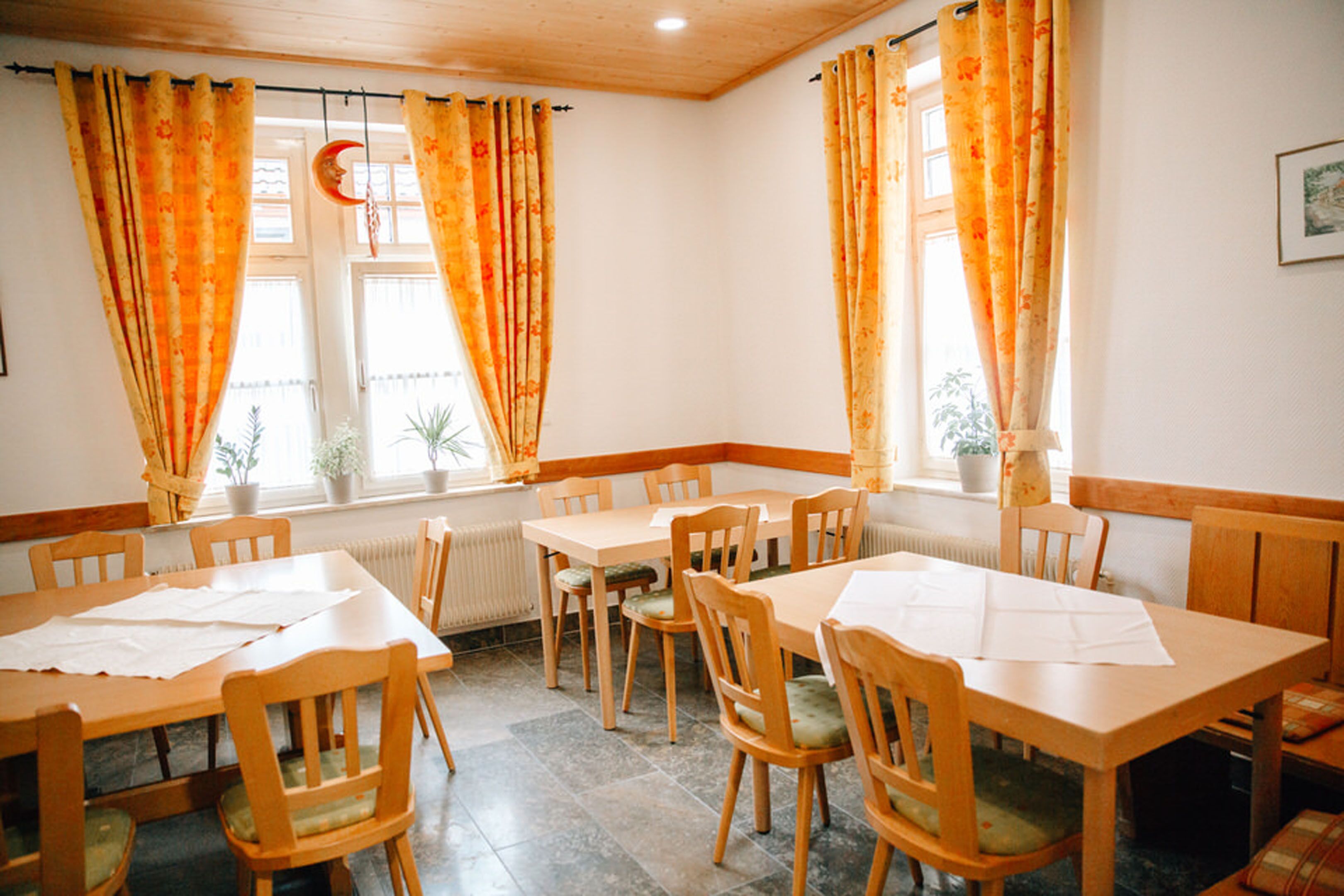 Uma área de refeições com mesas e cadeiras de madeira, cortinas laranja e plantas em vasos.