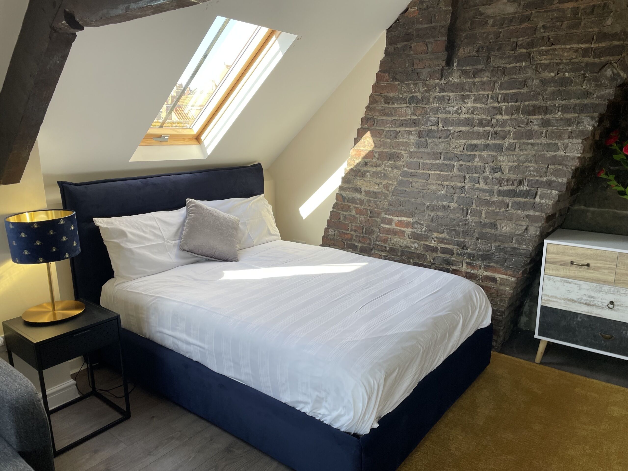 Um quarto com teto inclinado, um claraboia, uma cama com roupa de cama branca, uma cabeceira azul escura, uma mesa de cabeceira com abajur e uma cômoda com gavetas.