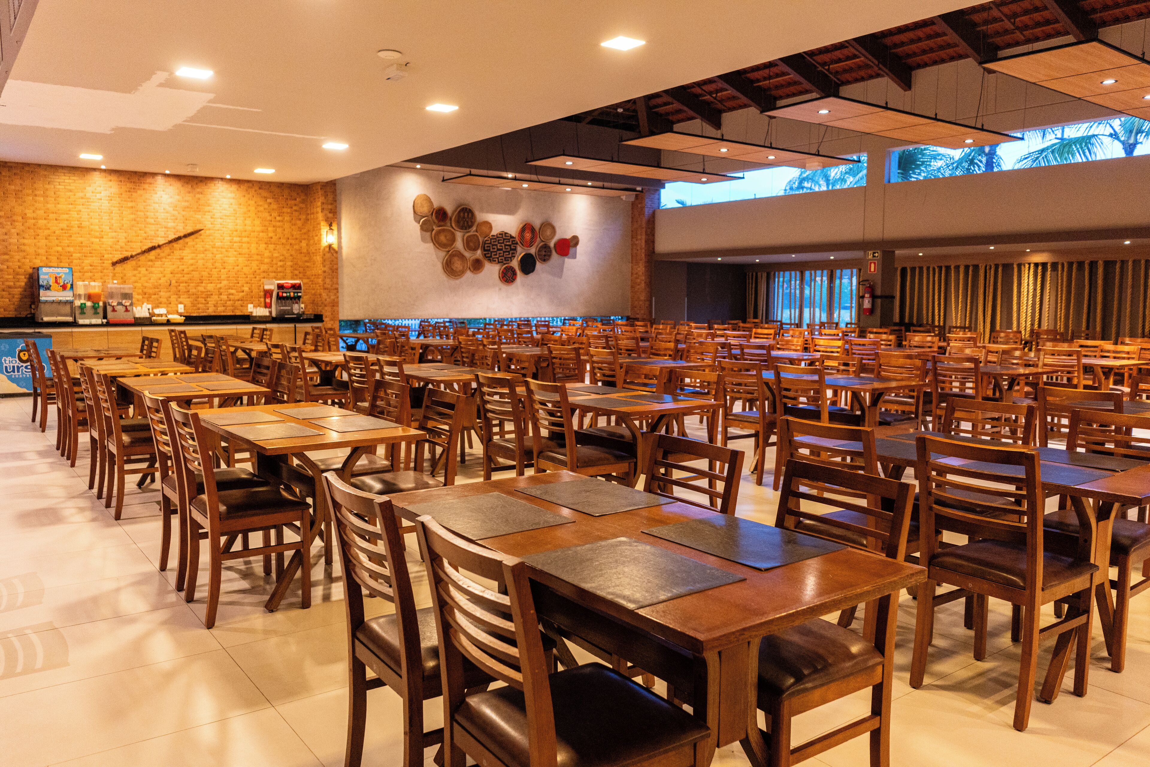 Uma ampla sala de jantar com várias mesas e cadeiras de madeira, uma parede de tijolos adornada com pratos decorativos, e uma máquina de café ao fundo.
