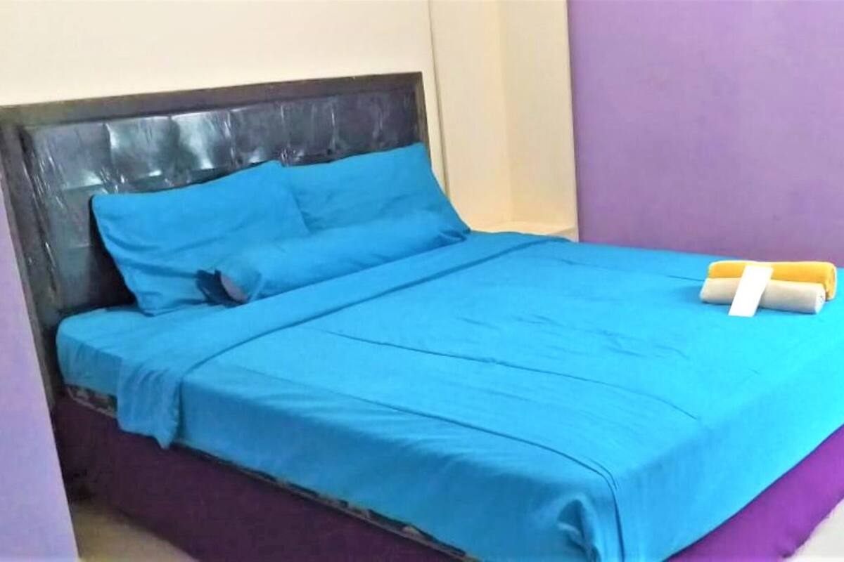 Uma cama com colcha azul e almofadas, cabeceira e uma toalha enrolada ao lado.