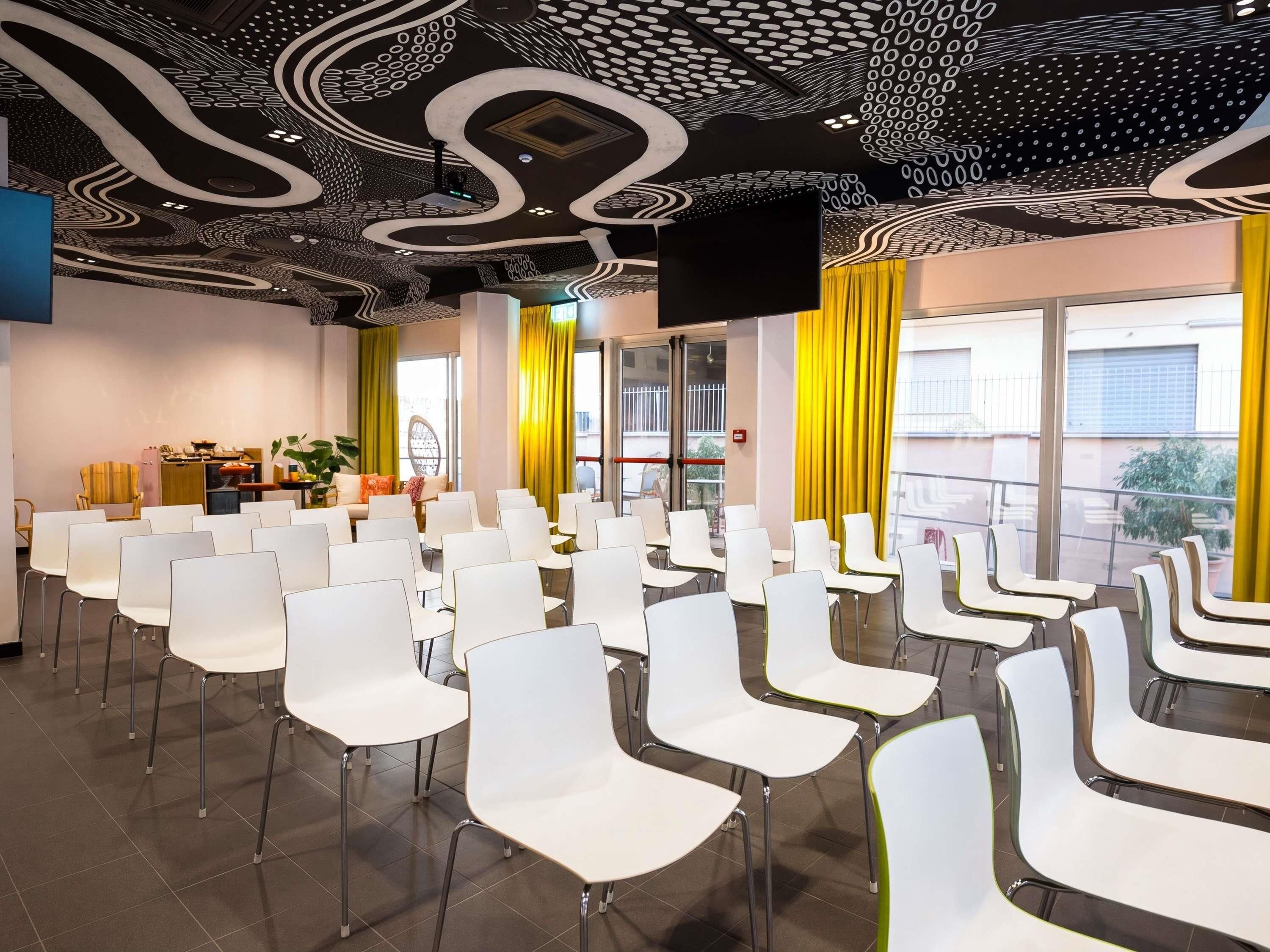 Uma sala de conferências com cadeiras brancas dispostas em fileiras, uma TV de tela plana instalada no teto e grandes janelas com cortinas amarelas.