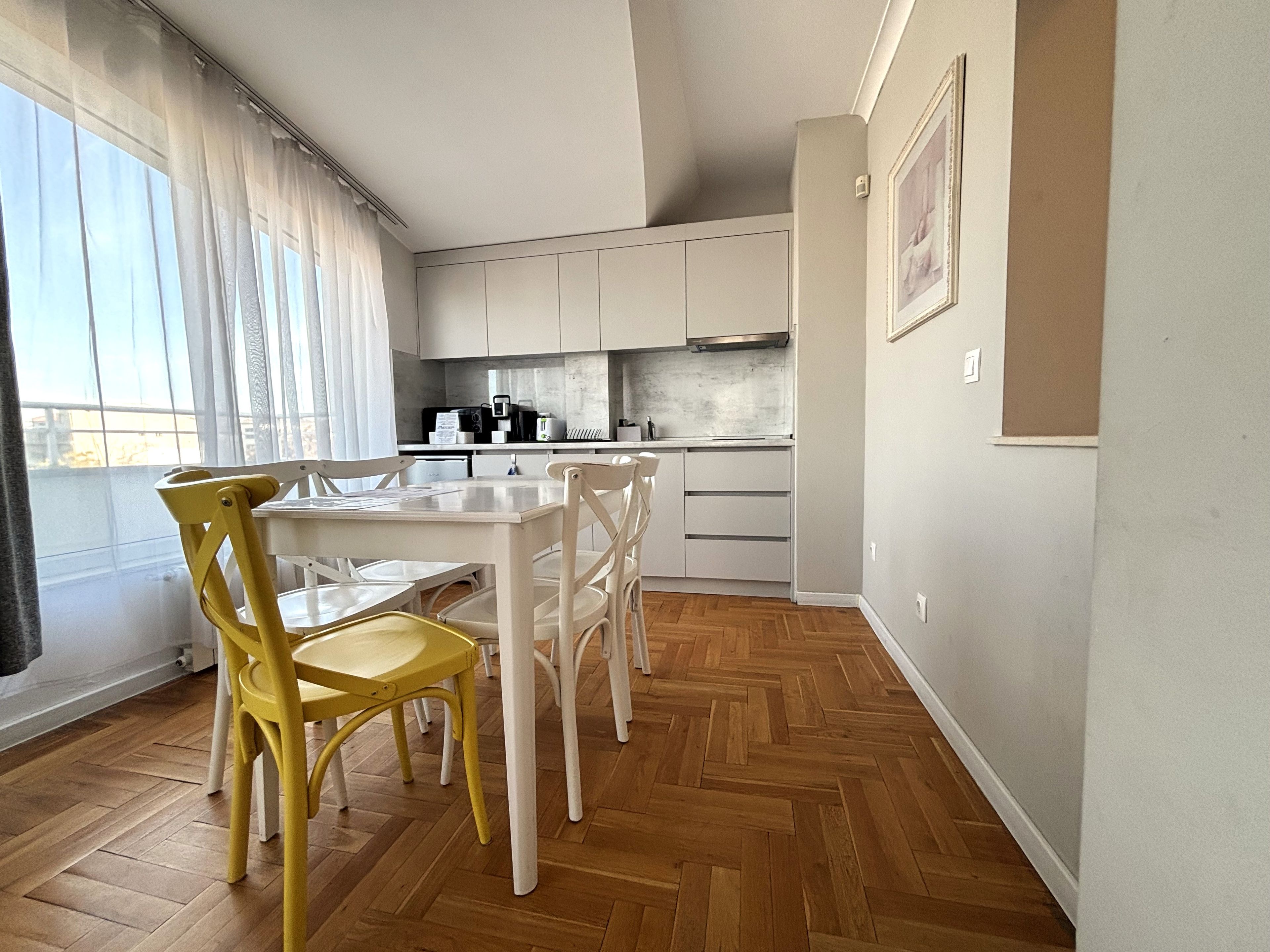 Cozinha compacta com armários brancos, uma mesa de jantar com cadeiras e piso de madeira em padrão herringbone.