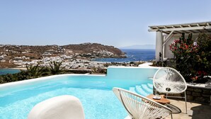 Piscina com vista para uma vila costeira, cadeiras brancas e uma mesa.