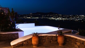Vista noturna de uma área de piscina com vasos de terracota e uma parede de pedra, com vista para um horizonte iluminado.