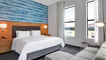 Quarto de hotel com uma cama grande, cabeceira de madeira, duas luminárias de cabeceira, um sofá cinza estofado e vista da cidade por grandes janelas.