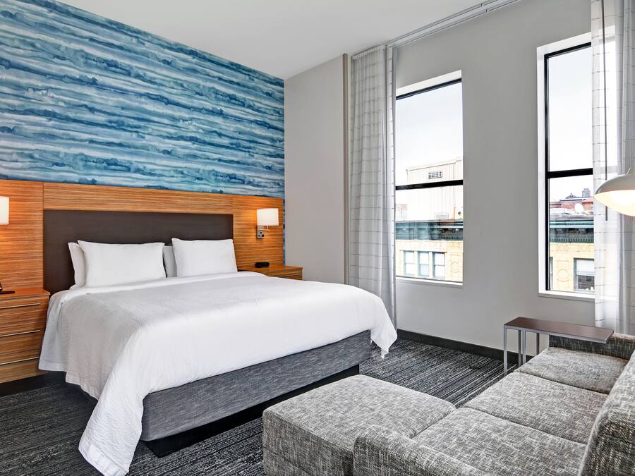 Quarto de hotel com uma cama grande, cabeceira de madeira, duas luminárias de cabeceira, um sofá cinza estofado e vista da cidade por grandes janelas.