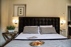 Quarto de hotel com uma cama grande, roupa de cama listrada, duas luminárias de cabeceira, um quadro com uma borboleta emoldurada, uma pequena planta em vaso e uma bandeja com croissants.