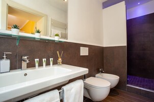 Uma casa de banho moderna com uma grande lavatório branco, duas sanitas, uma estante de vidro com plantas e uma área de duche.