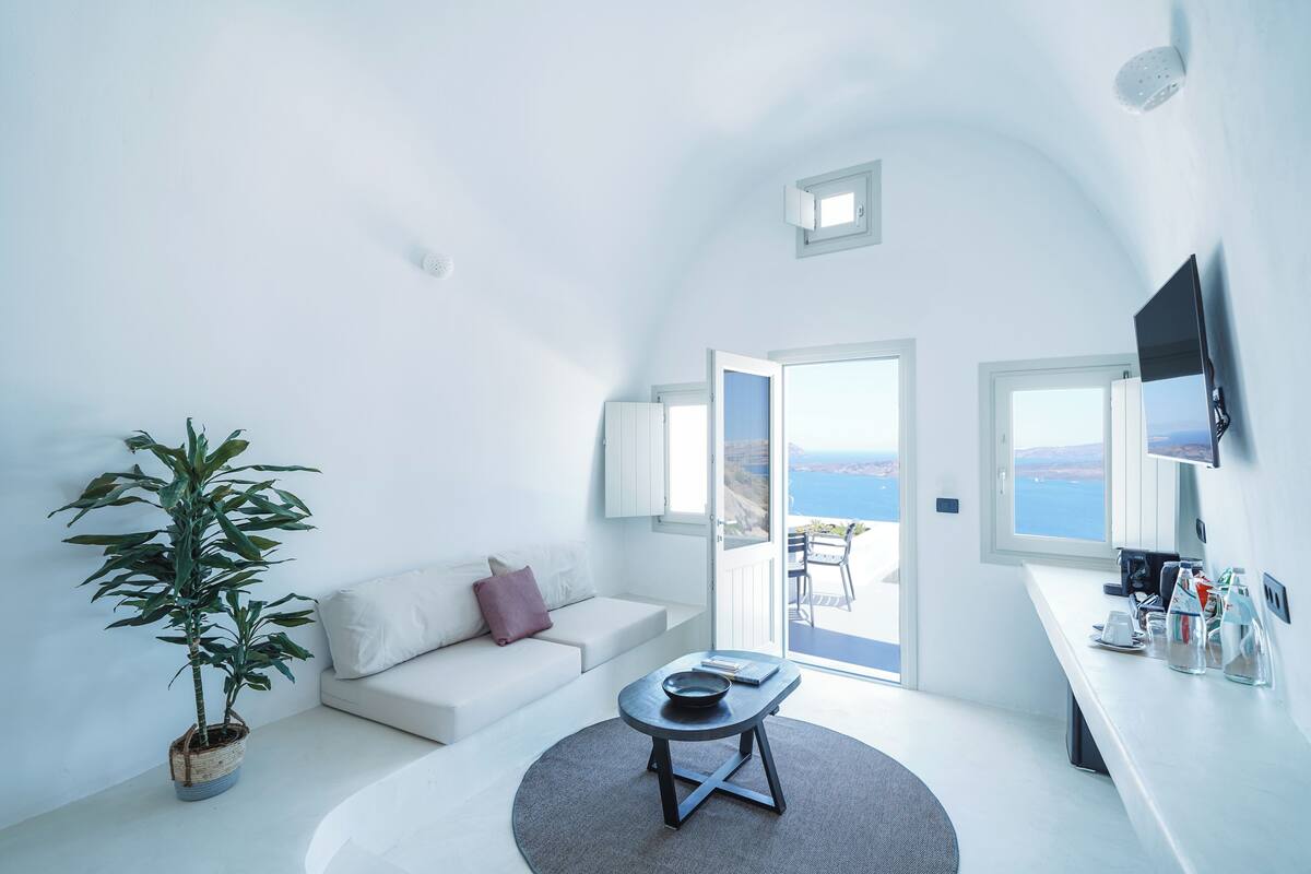 Quarto moderno com sofá branco, planta em vaso, mesa redonda e vista para o mar através de uma porta.