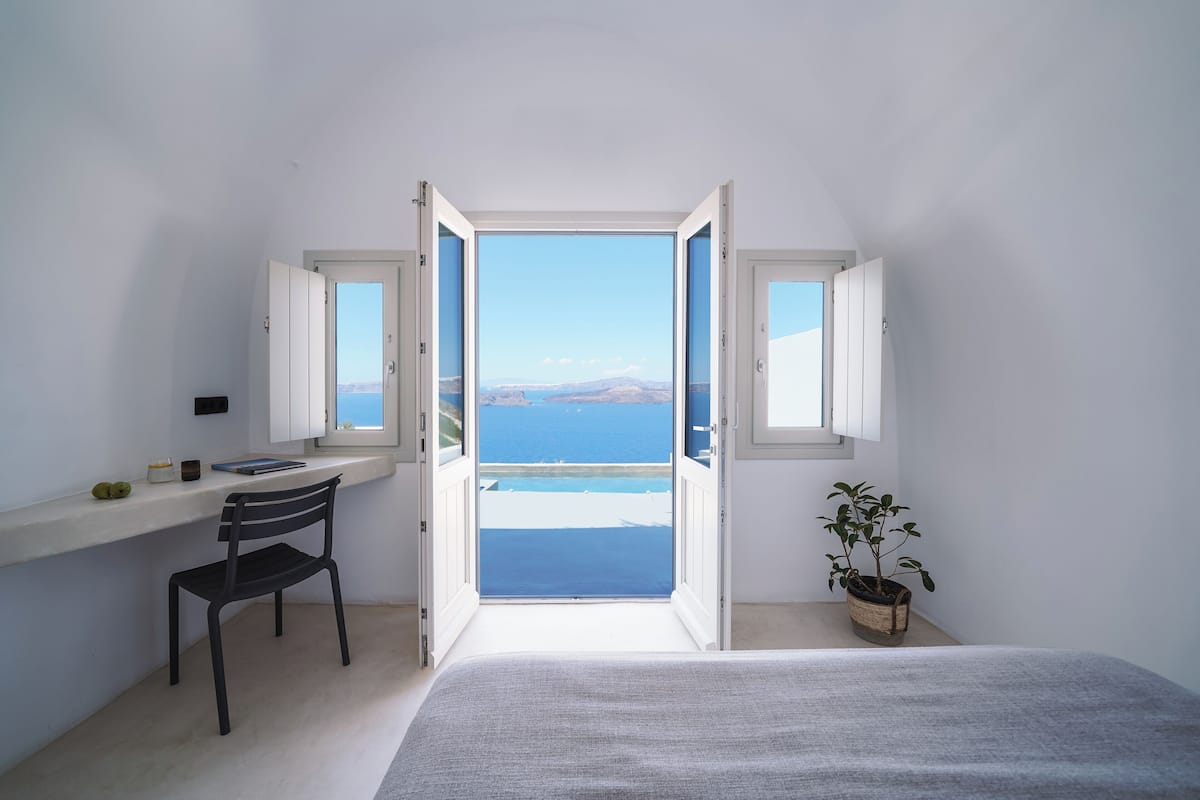 Quarto com cama, escrivaninha com cadeira, planta em vaso e vista para o mar através de uma porta aberta.