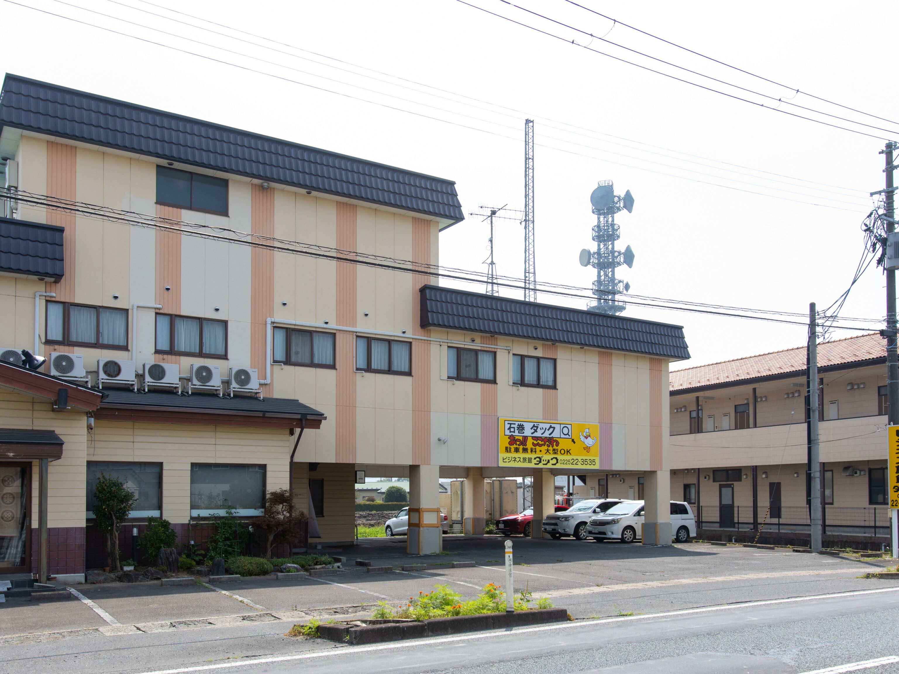 Um edifício de vários andares com fachada bege e marrom, vários aparelhos de ar condicionado no telhado, e um estacionamento com alguns carros.