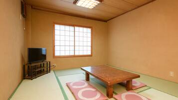Um quarto japonês tradicional com piso de tatame, uma mesa de madeira baixa, uma televisão em um suporte e uma grande janela com biombos shoji.