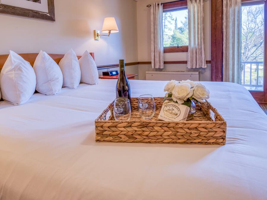 Cama bem arrumada com roupa de cama branca, uma garrafa de vinho, dois copos e um tabuleiro com rosas brancas.
