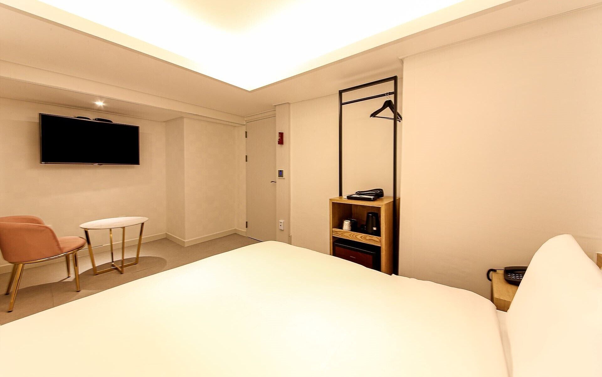Quarto de hotel com uma cama, televisão na parede, uma cadeira, uma mesinha e uma estante com chaleira e telefone.