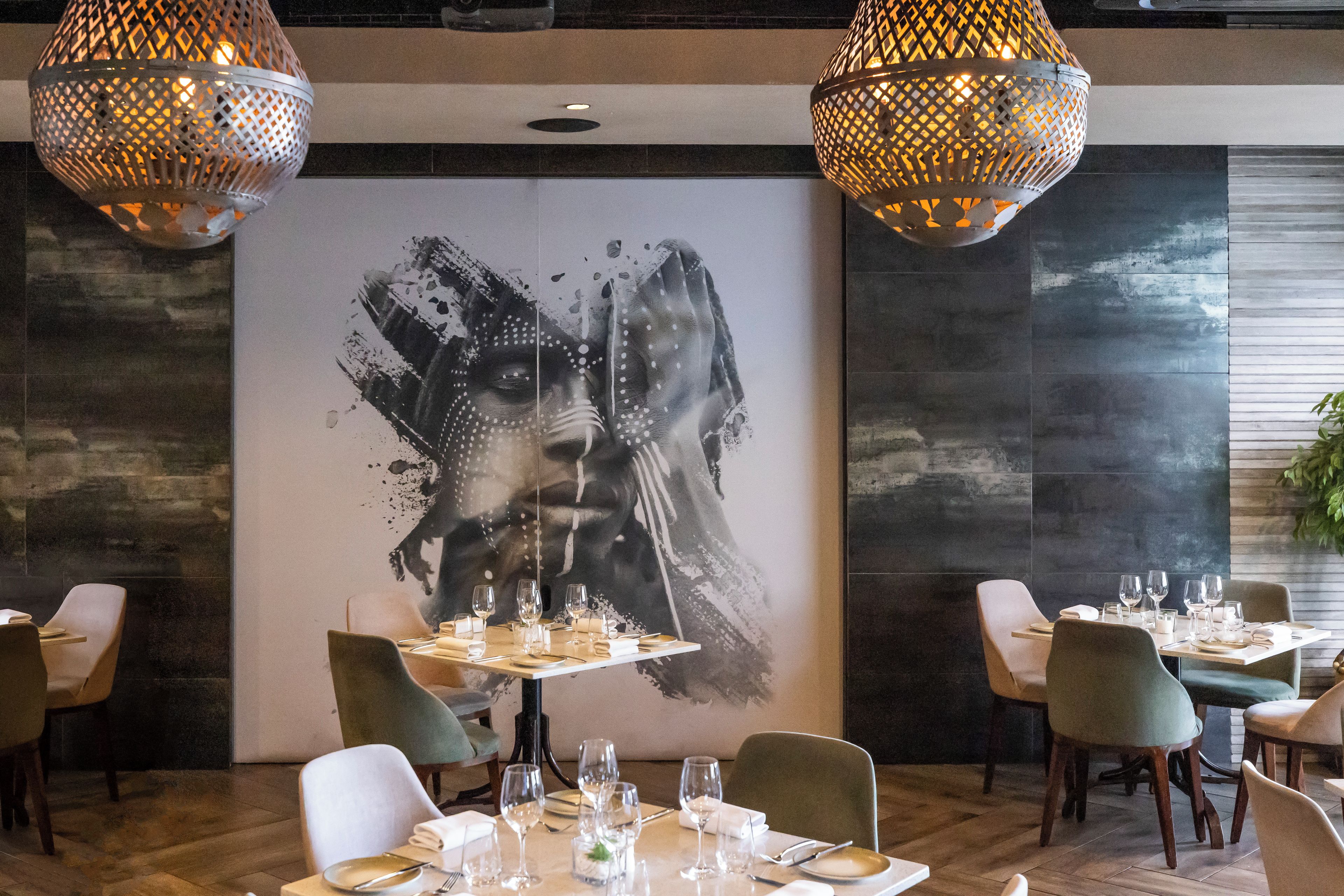 Interior de restaurante com uma grande pintura abstrata na parede, mesas preparadas para refeições e luminárias pendentes.