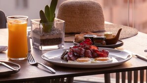 Uma mesa de jantar com um copo de suco de laranja, um pequeno vaso com planta, um chapéu de palha e um prato com comida.