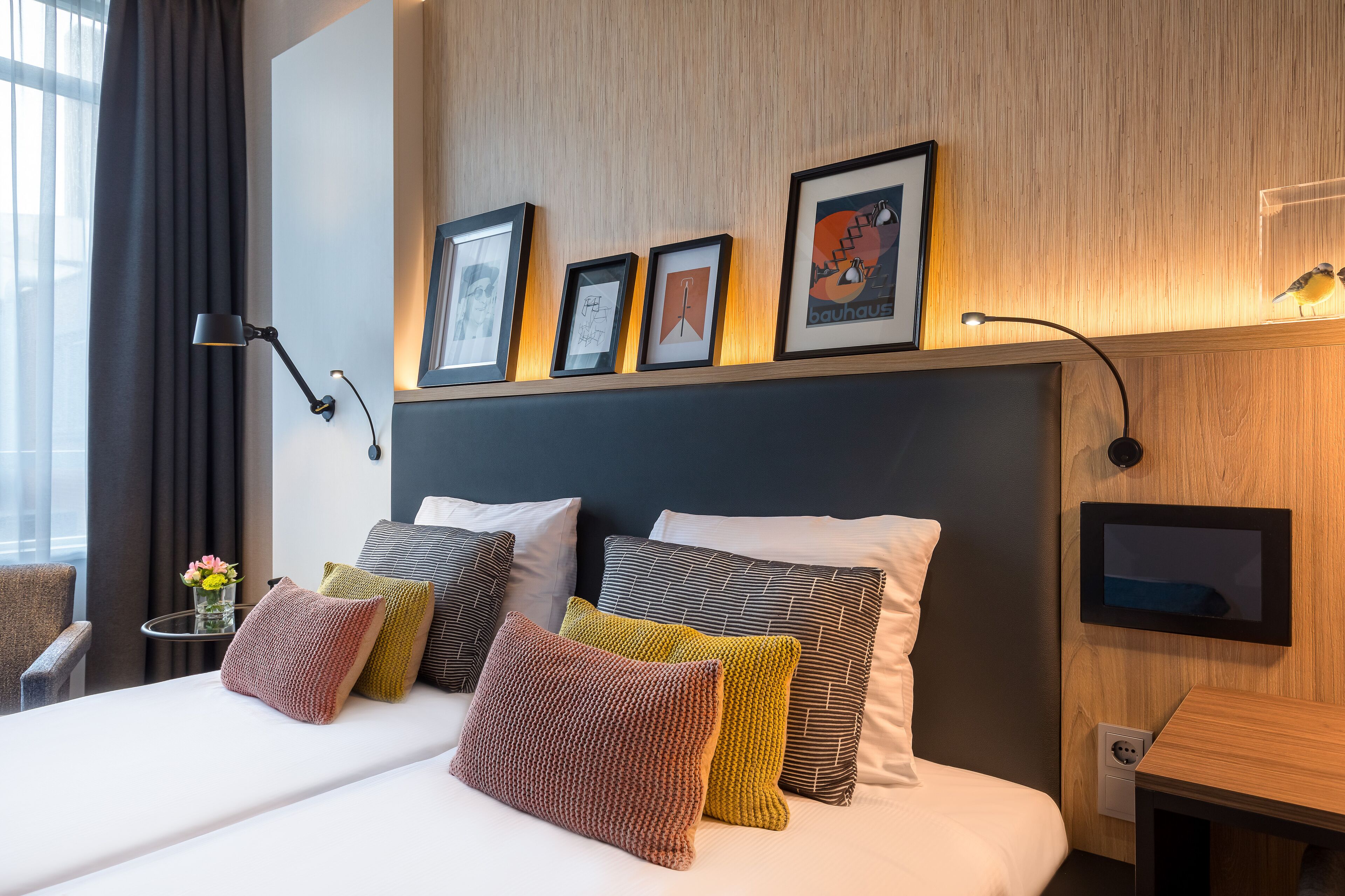 Quarto de hotel com uma cama bem arrumada, uma mesa de cabeceira com abajur, uma pequena mesa com um vaso de flores e obras de arte emolduradas na parede.