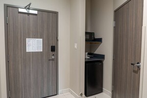 Quarto com duas portas de madeira, uma delas com a placa do plano de emergência, uma pequena geladeira preta e uma prateleira acima.